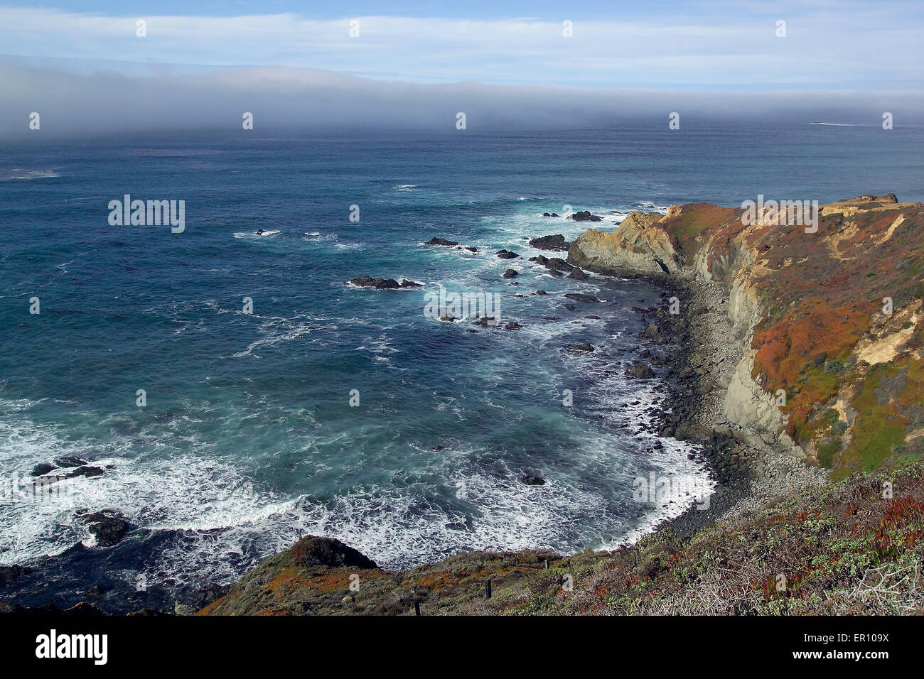 A gray fog bank rolls in over the blue Pacific Ocean along the rugged ...