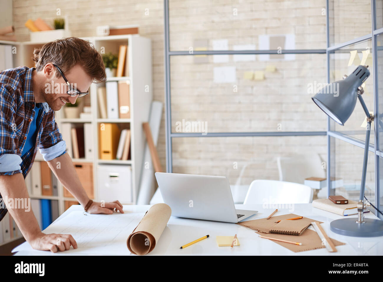 Happy architect looking at blueprint by workplace Stock Photo - Alamy