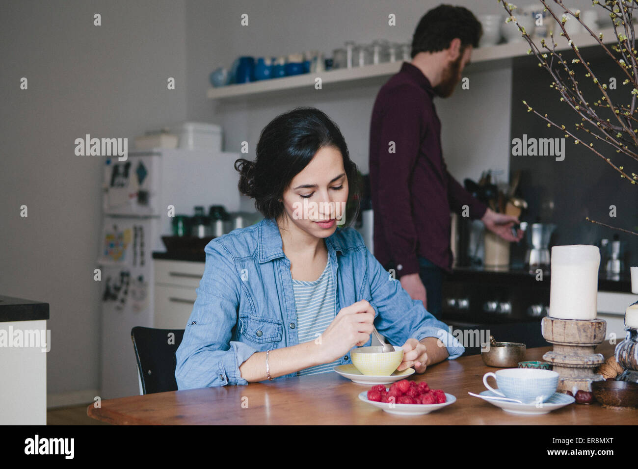 Young woman stirring coffee at kitchen table with man in background ...