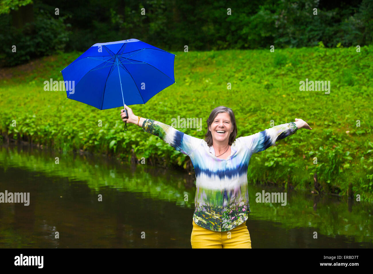 Older Woman Standing By A River With A Blue Umbrella And Spreading Her