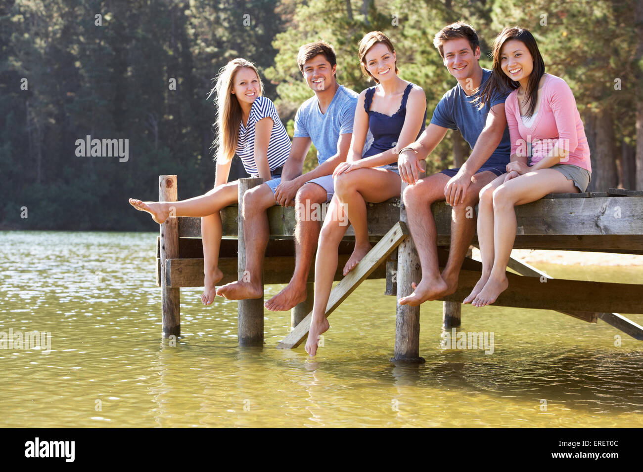 Group Of Young Friends Sitting On Wooden Jetty Looking Out Over Lake ...