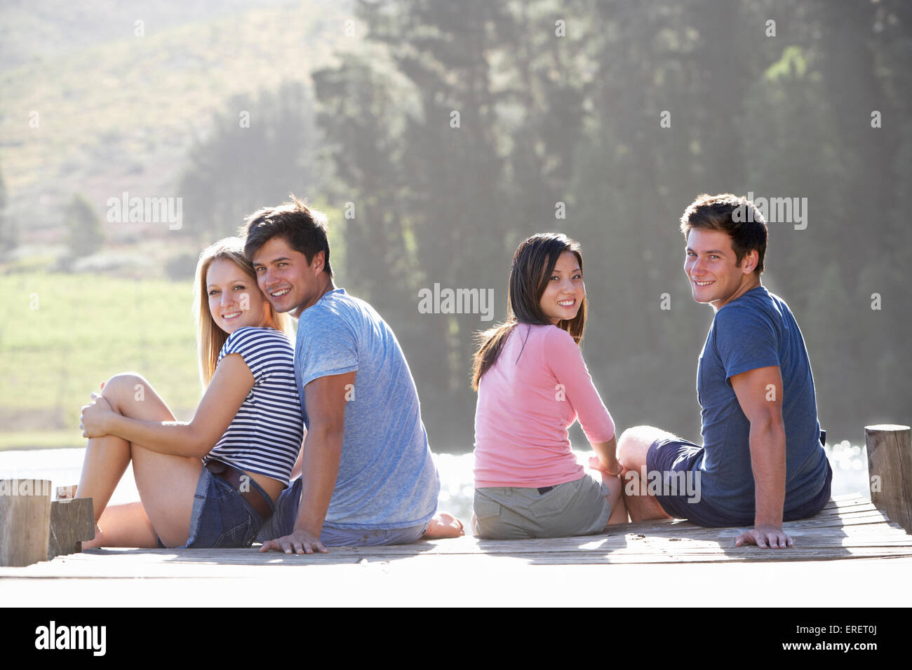Group Of Young Friends Sitting On Wooden Jetty Looking Out Over Lake ...