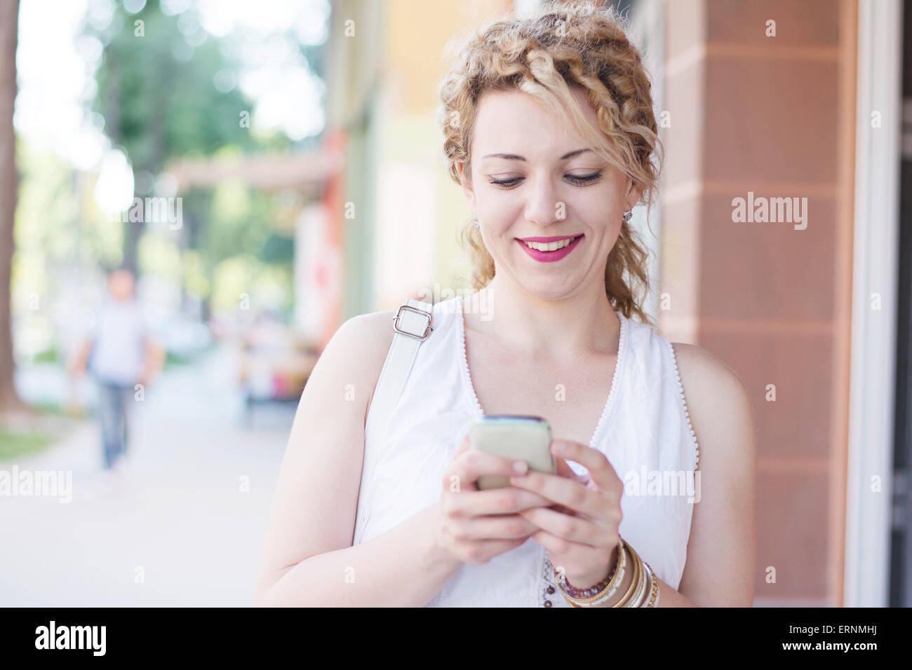 Curly blond girl typing on her phone on the street. Smiling while ...