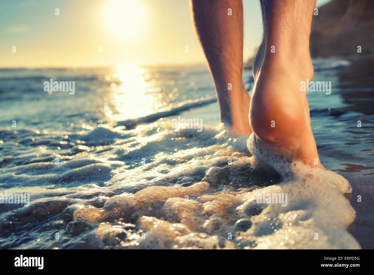 Closeup of a man's bare feet walking at a beach at sunset, with a wave's edge foaming gently ...