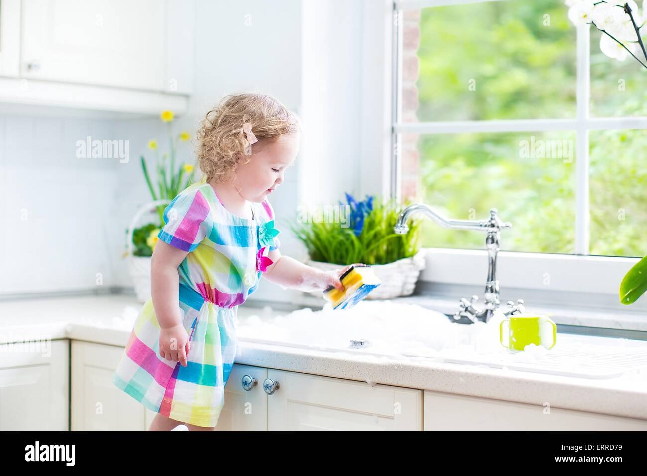 Cute curly toddler girl in colorful dress washing dishes, cleaning with ...