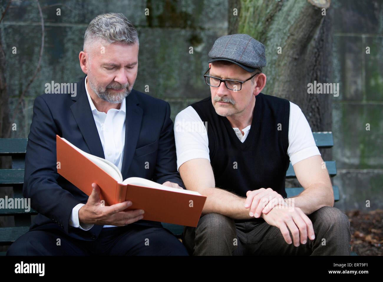 two men sitting outdoors on a bench with a book Stock Photo - Alamy
