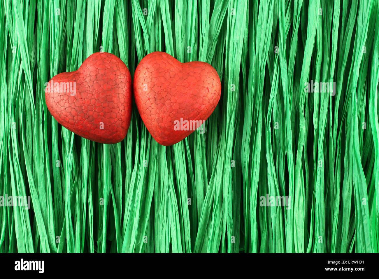 Two red painted polyfoam hearts on green paper raffia strips background ...