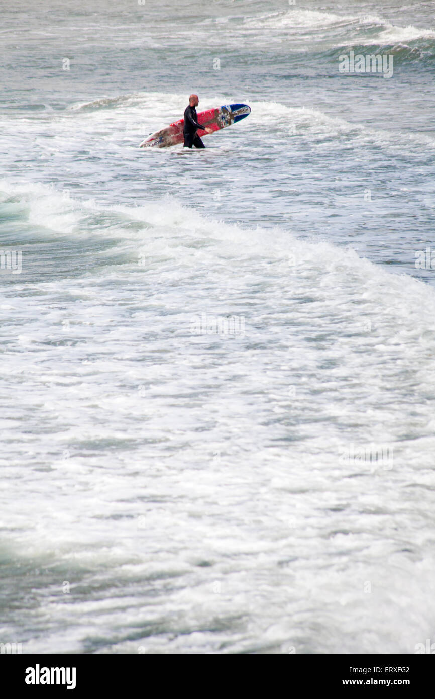 Bournemouth, Dorset, UK 9 June 2015. Surfer walking in the sea holding ...