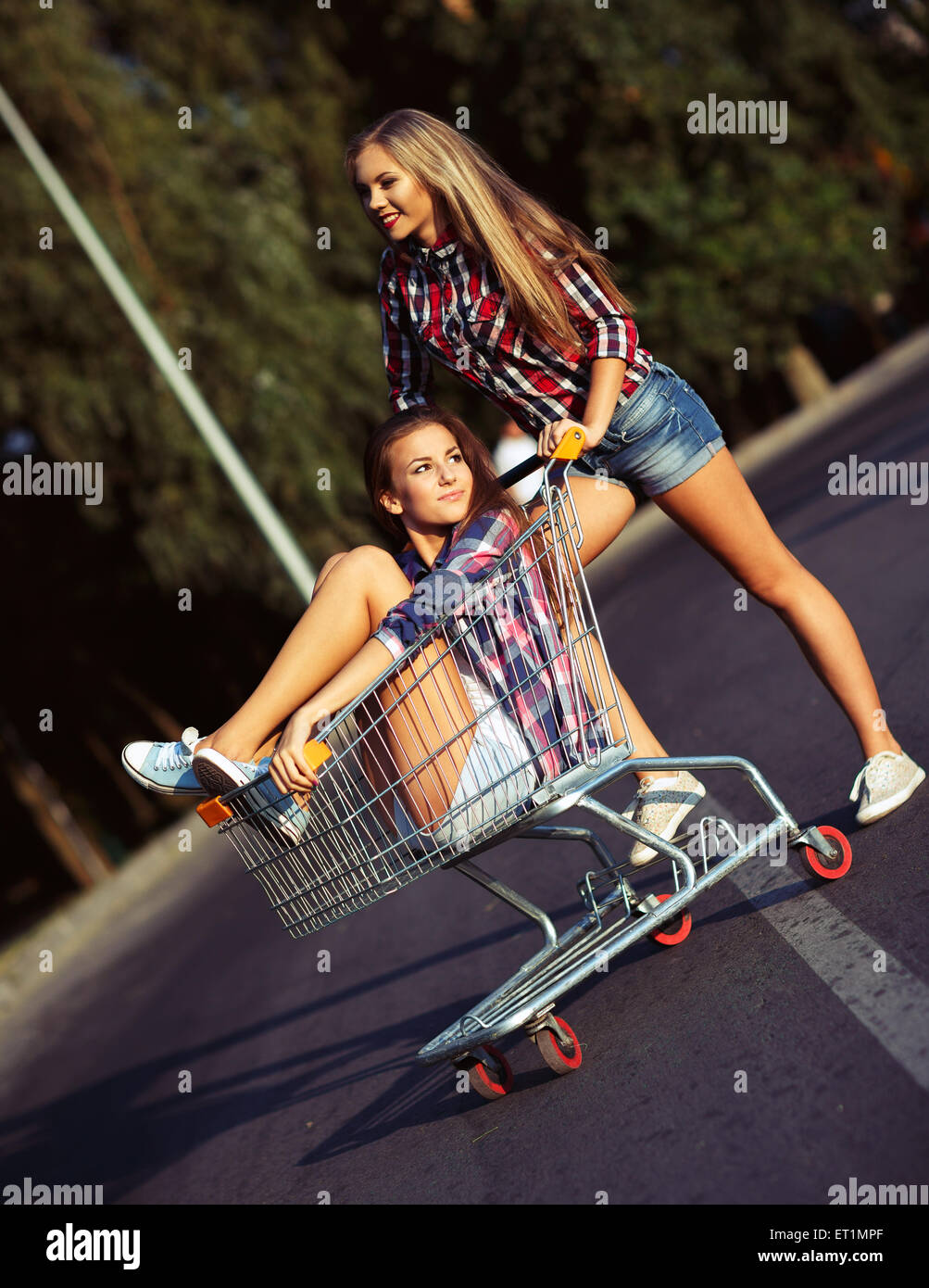 Two happy beautiful teen girls driving shopping cart outdoors ...