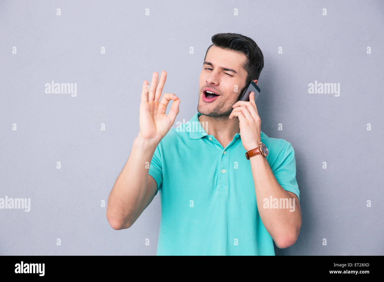 Happy young man talking on the phone and gesturing ok sign over gray ...