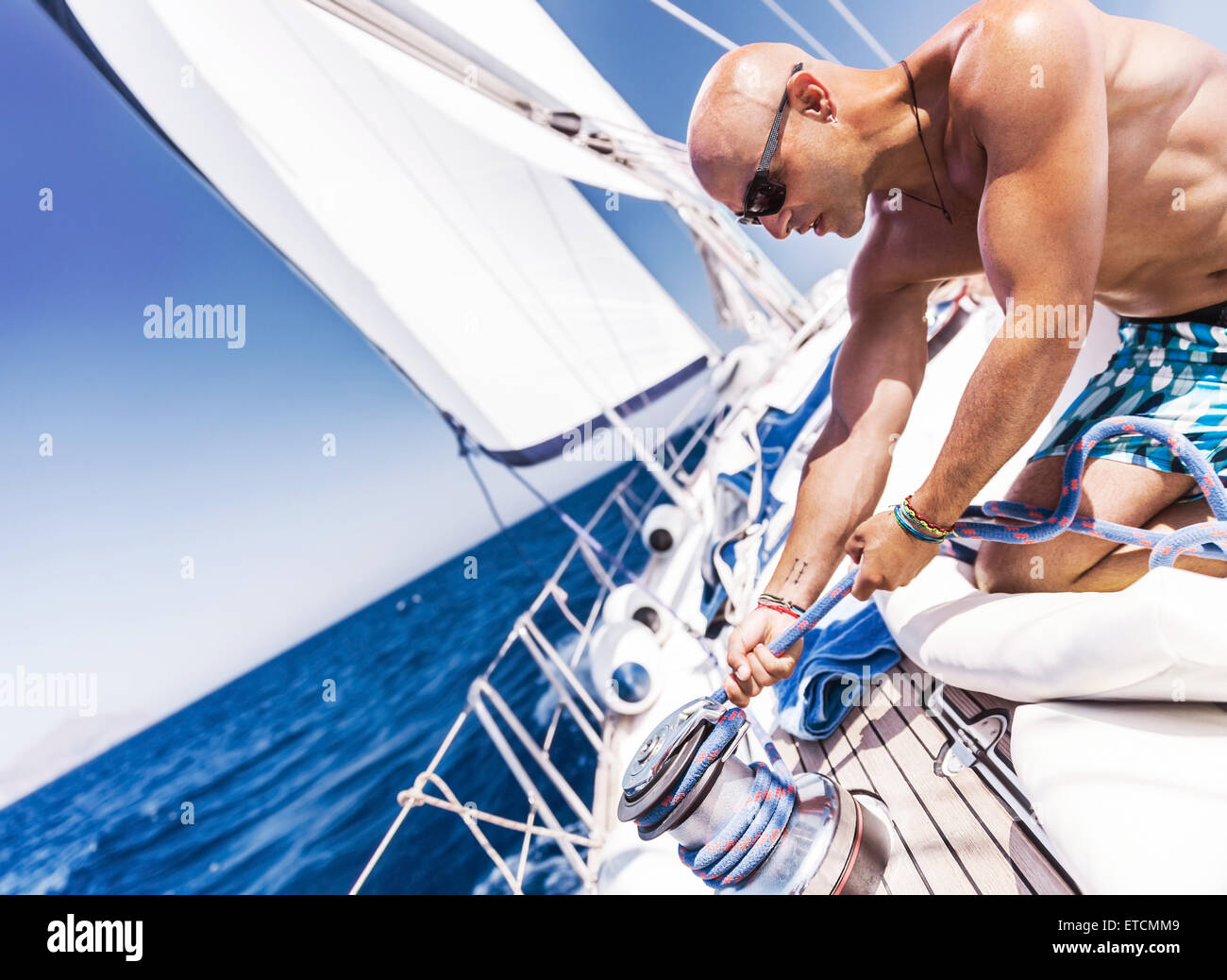 Handsome shirtless sailor working on sailboat, pulling rope on crank ...