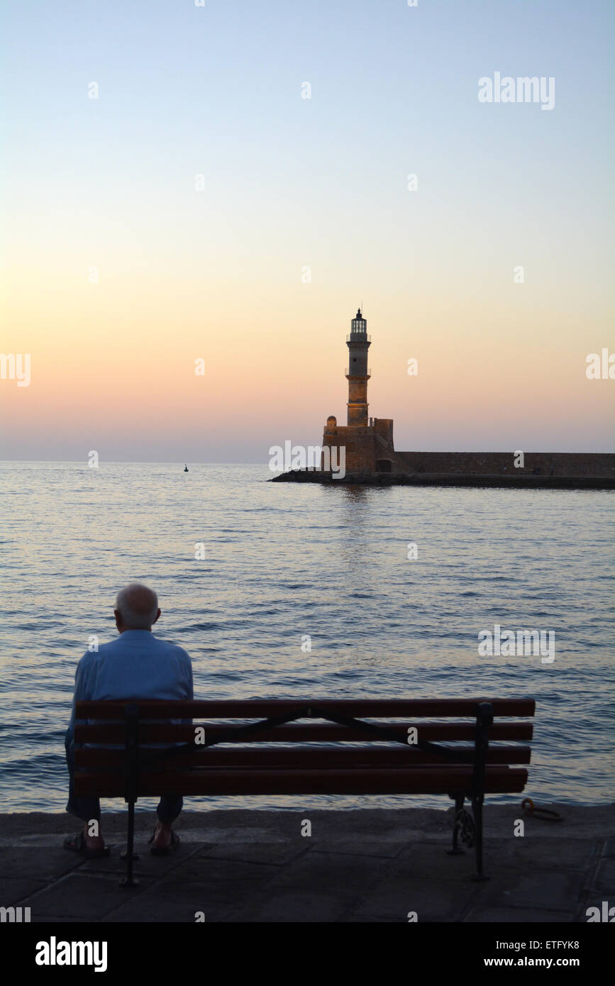A man sitting alone on a bench looking out to sea and the lighthouse at ...