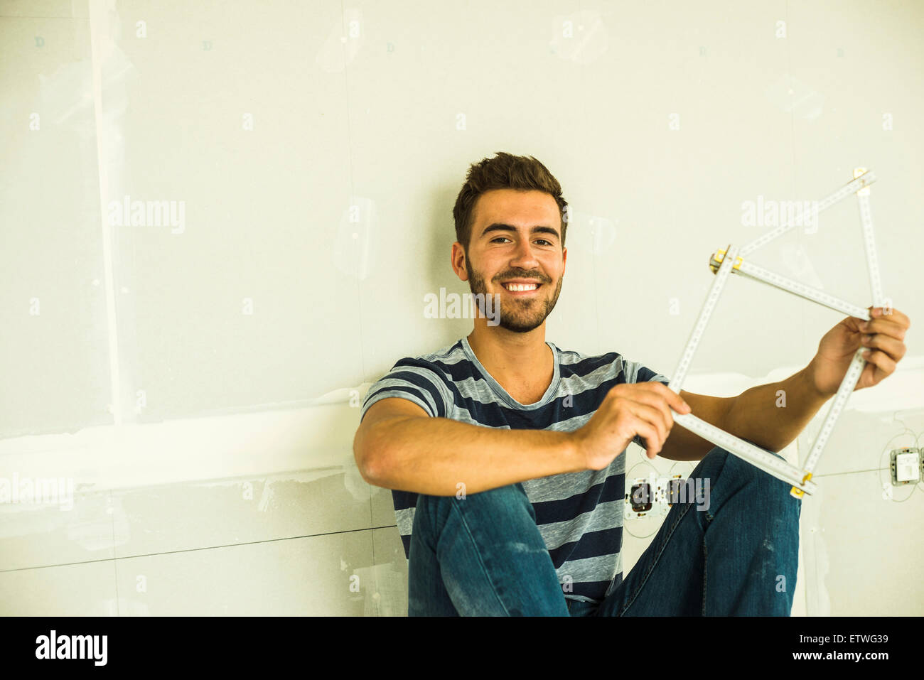 Smiling young man sitting at blank wall holding pocket rule Stock Photo ...