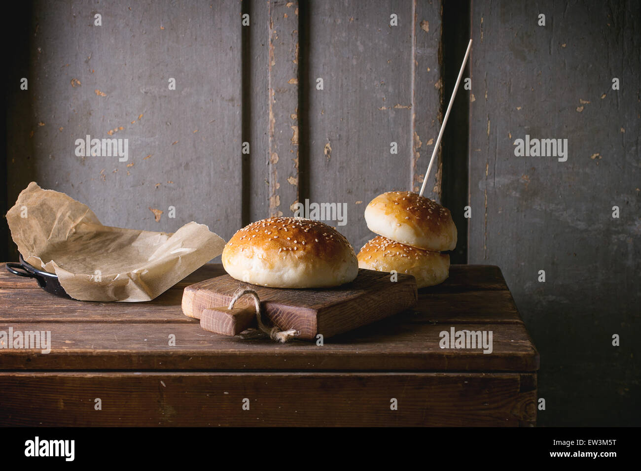 Fresh baked burger buns on small cutting board over wooden table. Dark ...