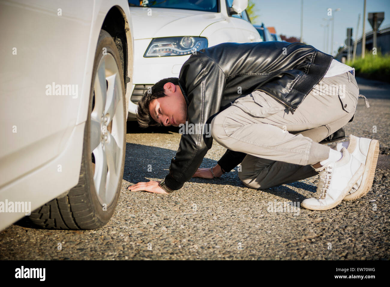 Concerned Young Man Kneeling on Pavement and Examining Underside of Car ...