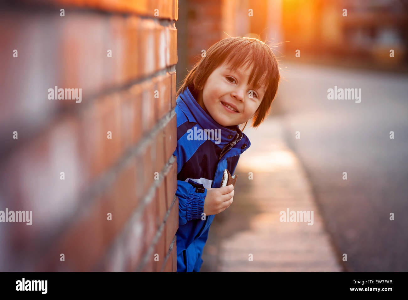 Adorable little boy, next to brick wall, eating chocolate bar on sunset ...