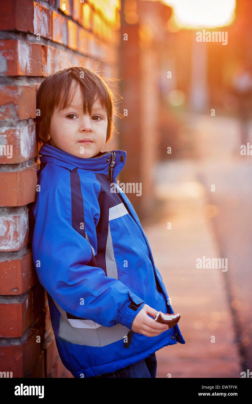 Adorable little boy, next to brick wall, eating chocolate bar on sunset ...