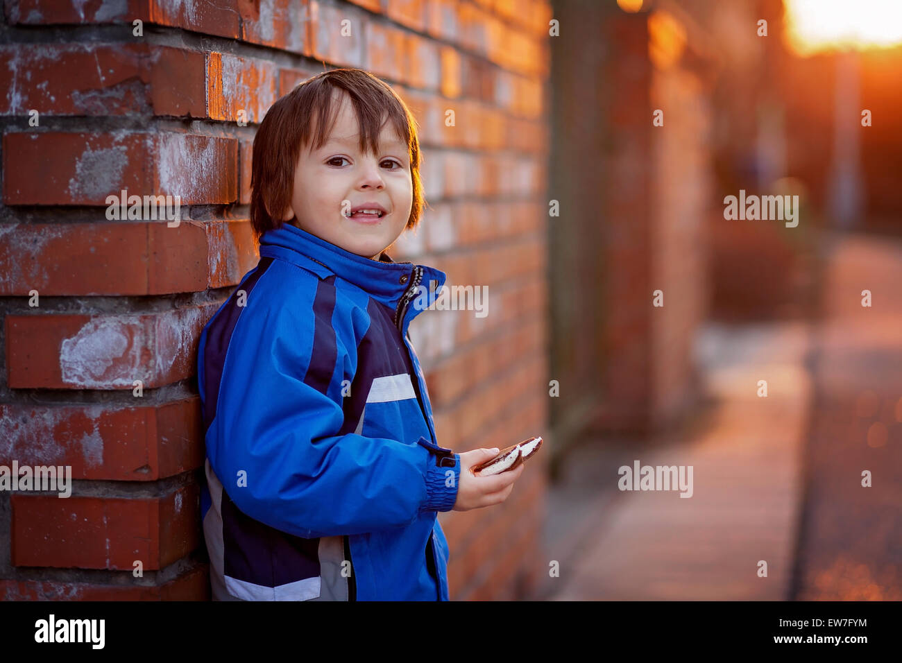 Adorable little boy, next to brick wall, eating chocolate bar on sunset ...
