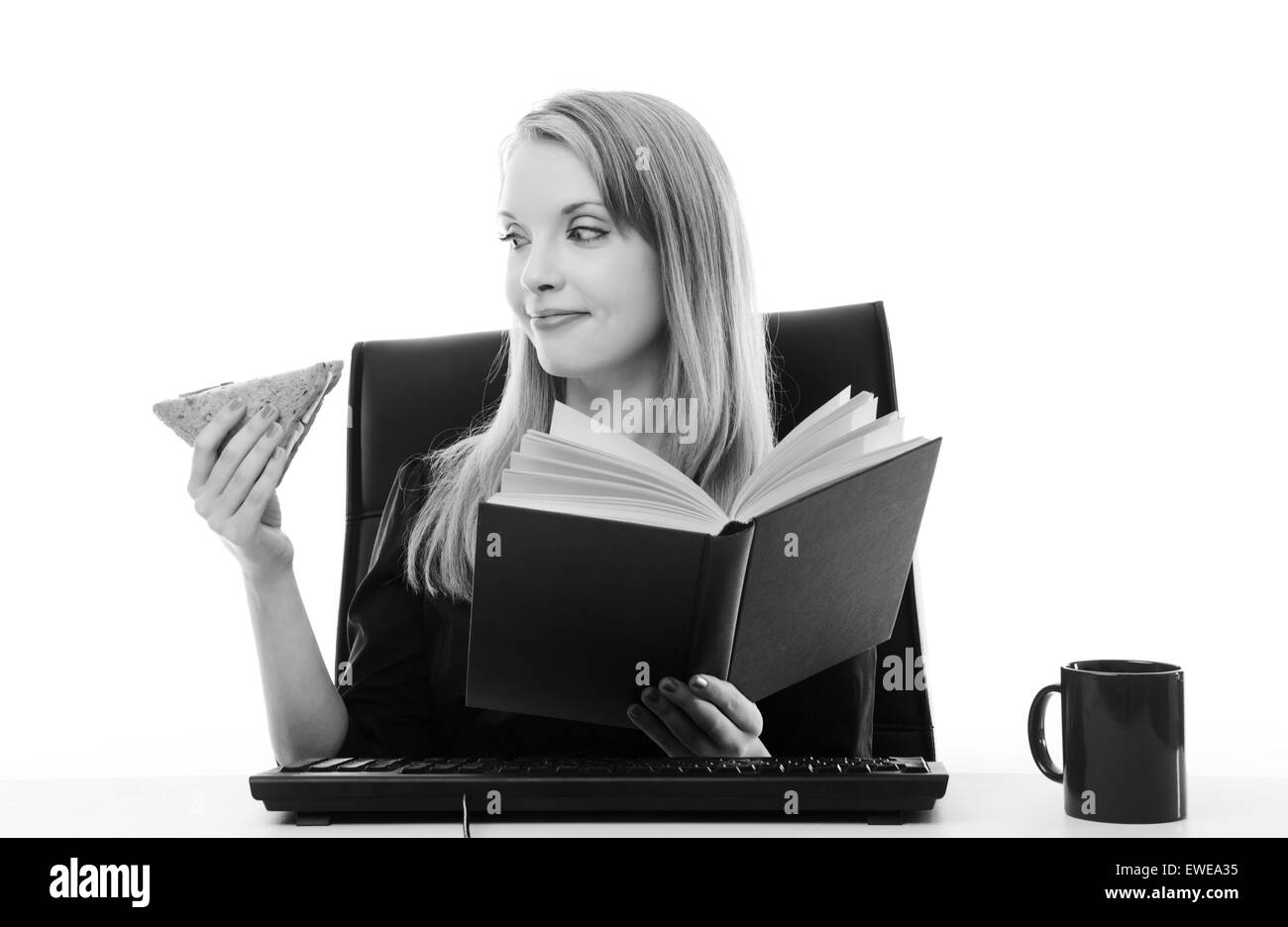 woman at work sitting at her desk reading a book and eating a sandwich ...