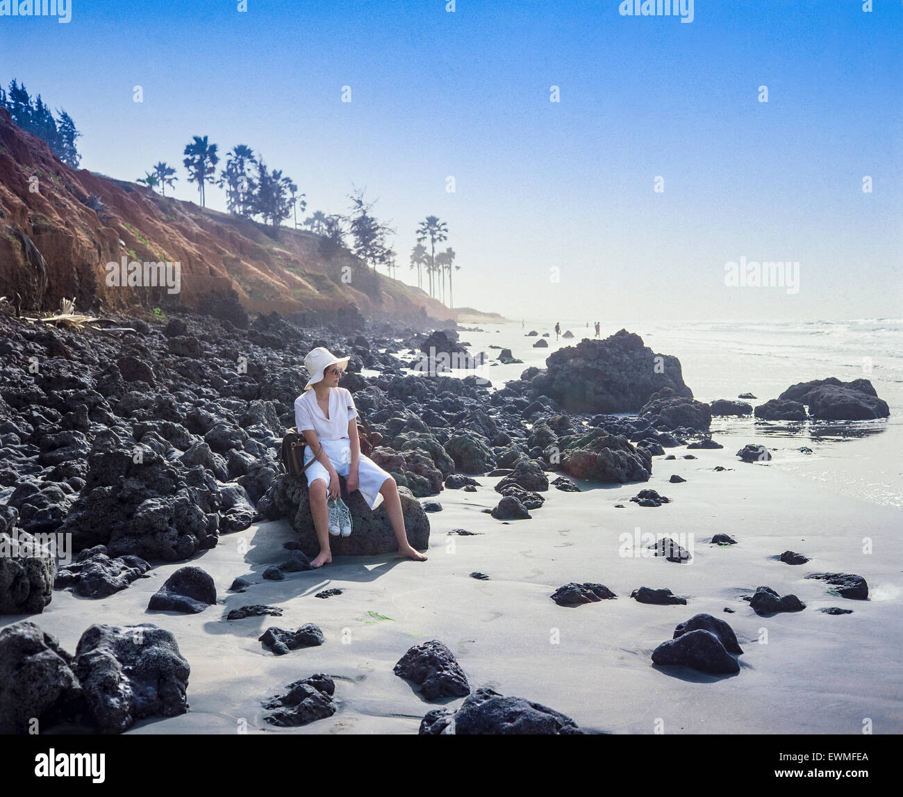 Woman with white dress and hat sitting on rock, Fajara beach, Gambia ...