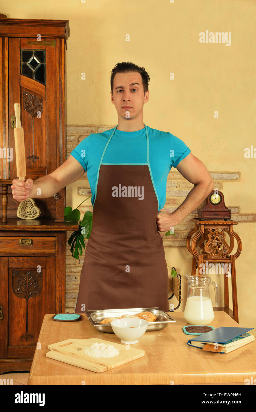 young man in an apron holding rolling pin while standing in kitchen ...