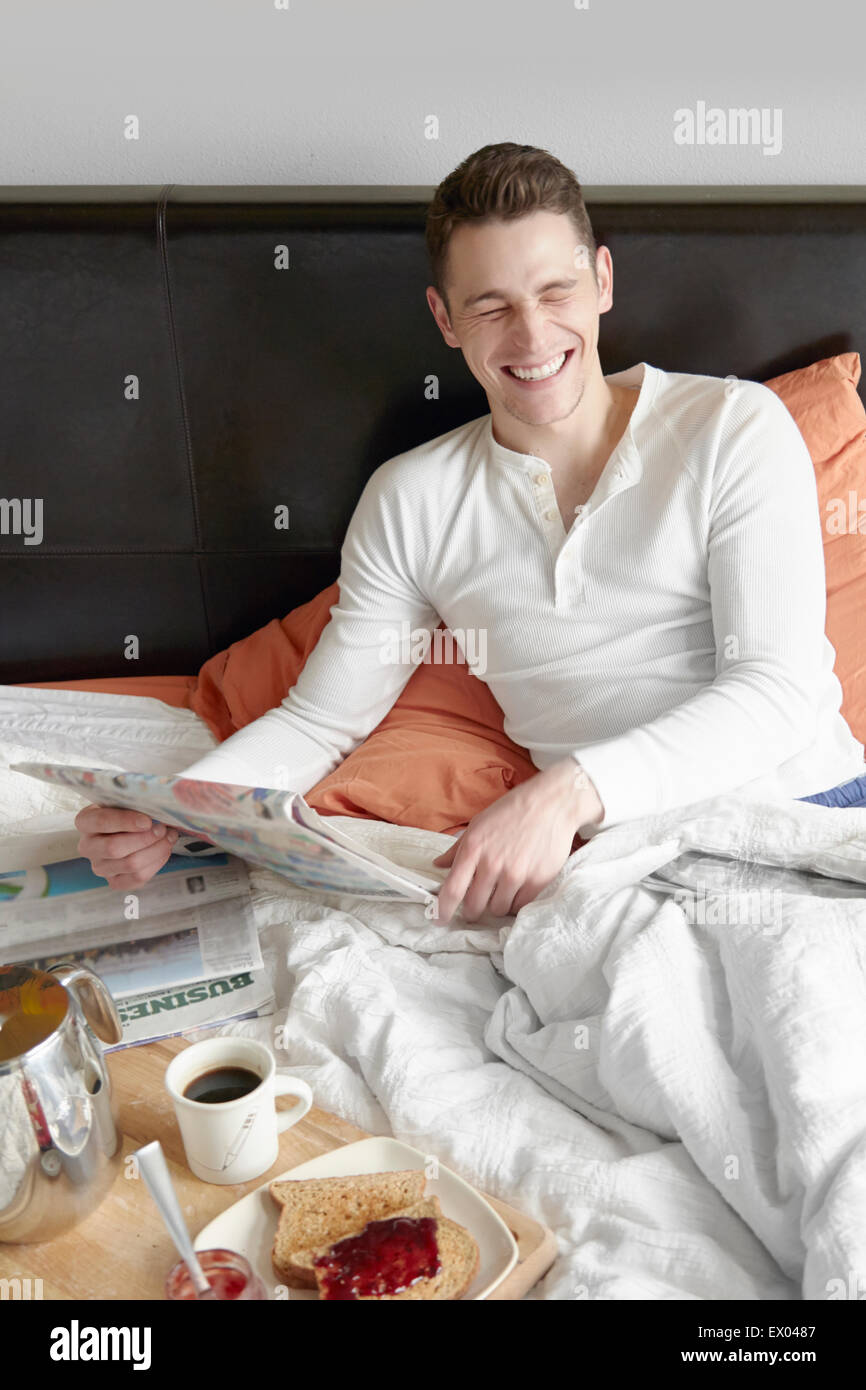 Young man sitting up in bed and laughing whilst reading newspaper Stock ...