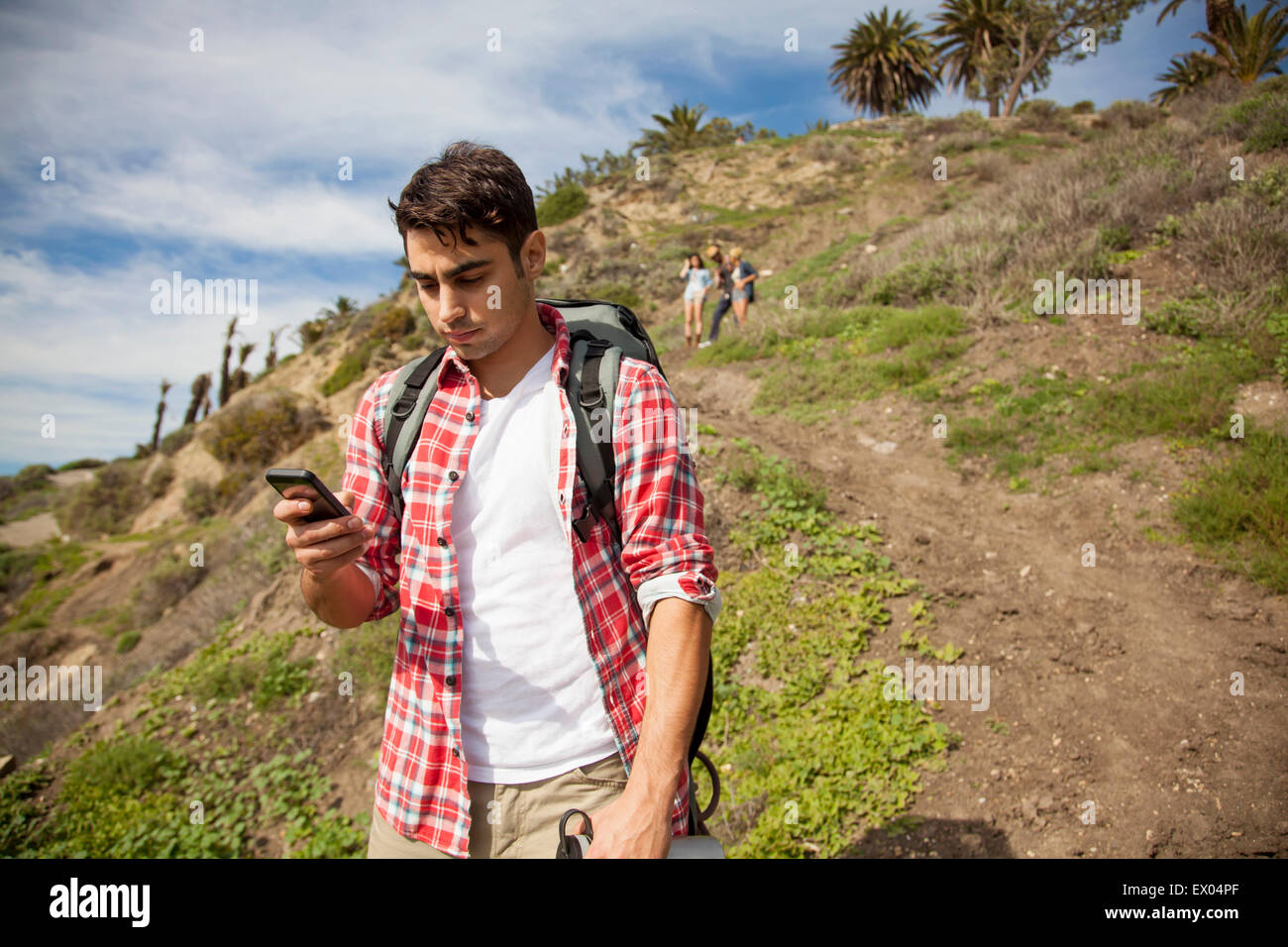 Young man at bottom of hill, using smartphone, friends trailing behind ...