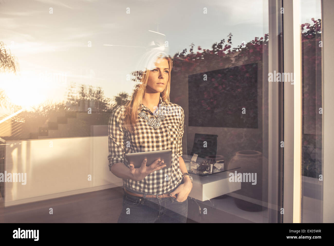 Portrait of young woman with digital tablet gazing from suburban window ...