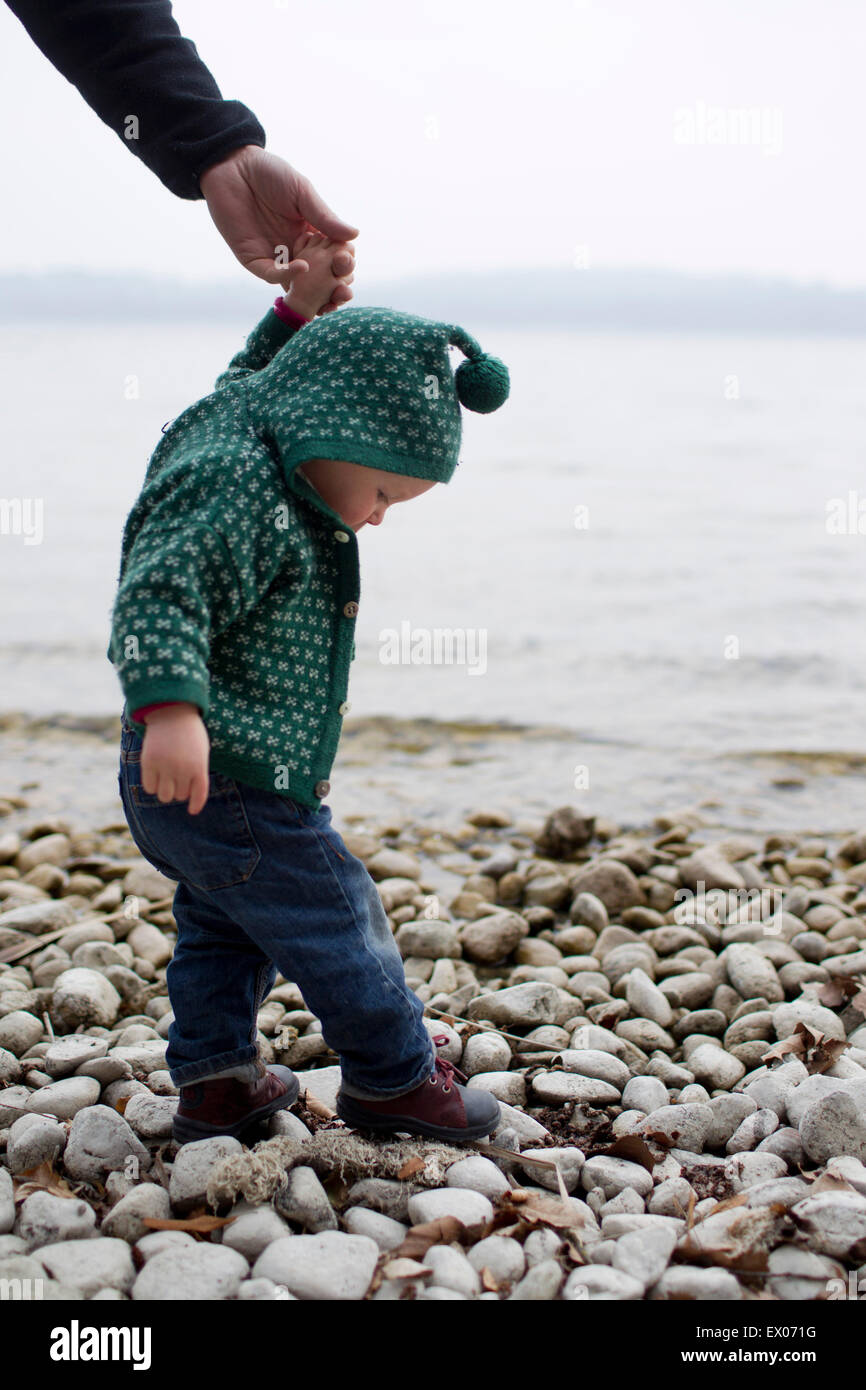 Baby girl holding fathers hand stepping on lakeside pebbles, Lake ...