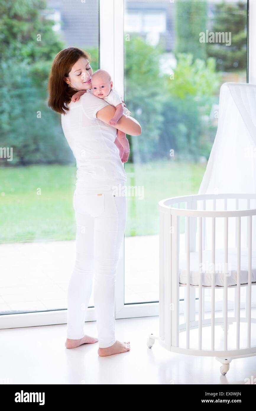 Young happy mother holding her newborn baby standing next to a window ...