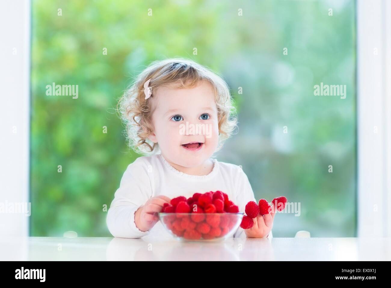 Beautiful little girl eating raspberry in a white dining room at a ...
