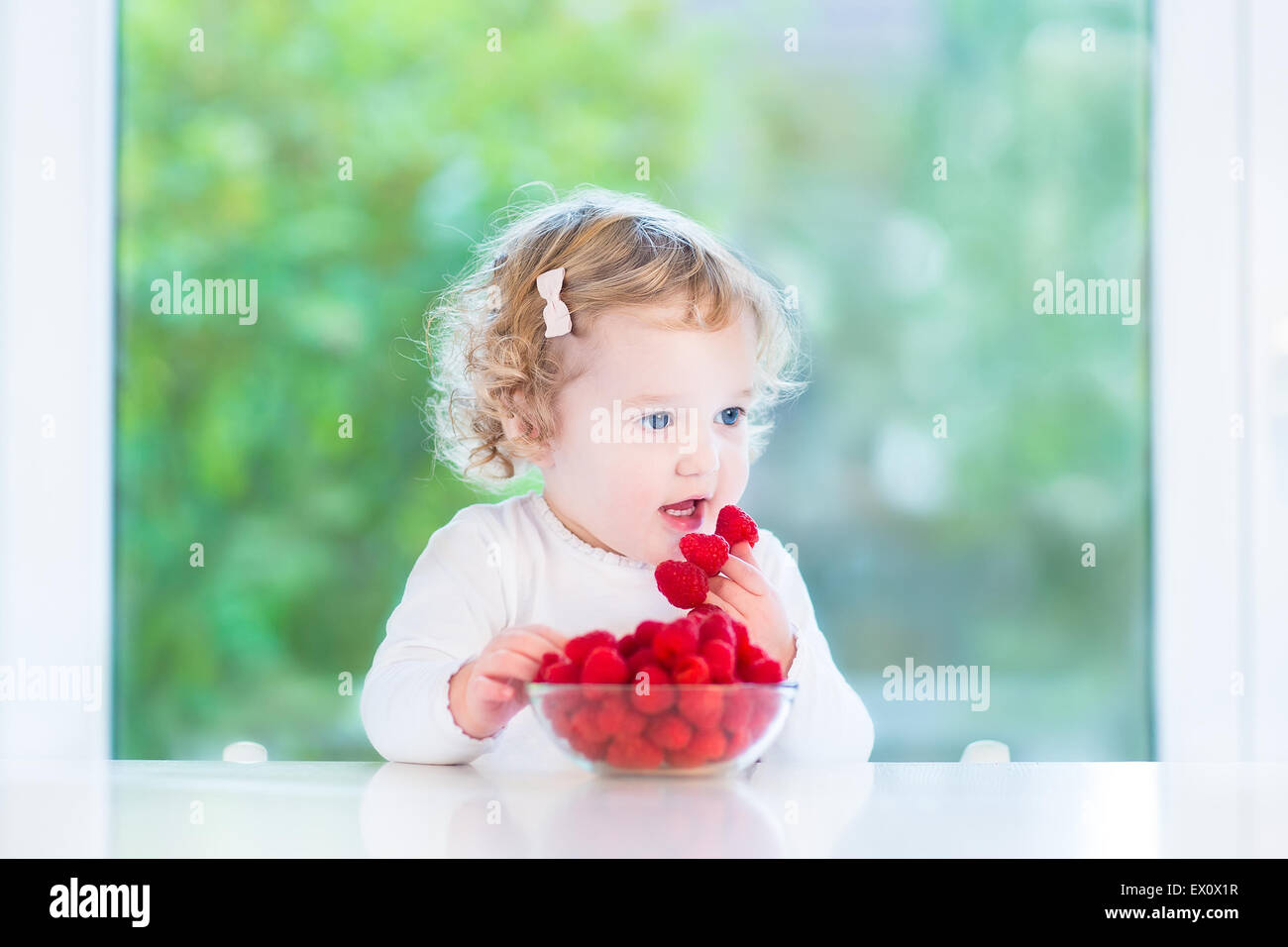 Adorable baby girl eating raspberry at a white table next to a big ...