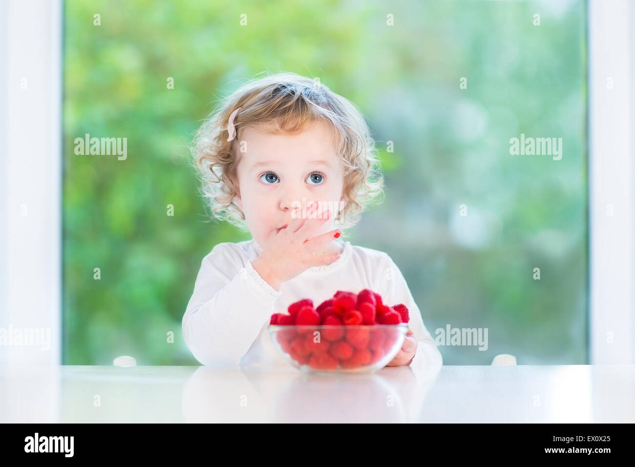 Adorable baby girl eating raspberry at a white table next to a big ...