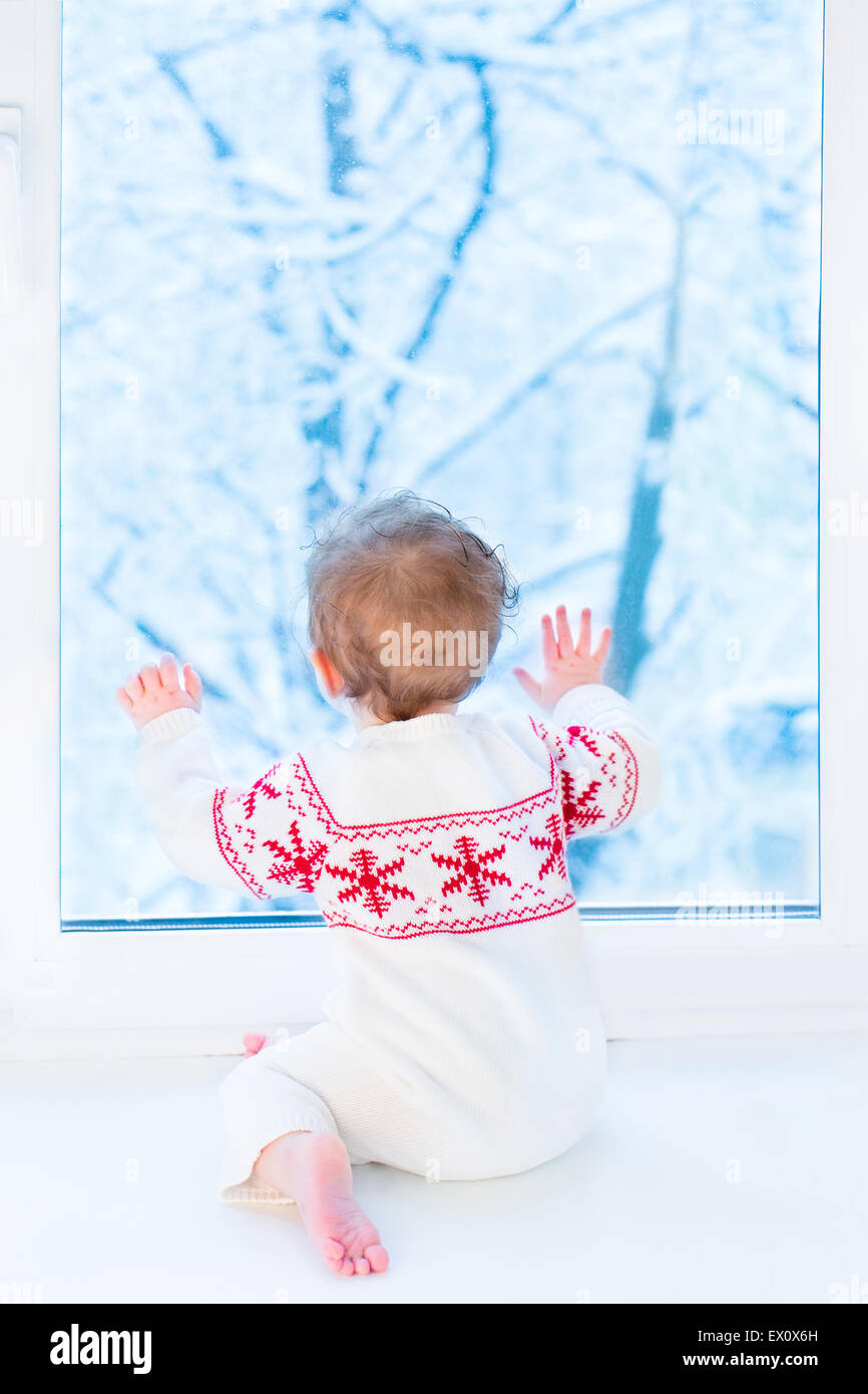 Happy smiling baby sitting at a window watching snow covered trees in ...