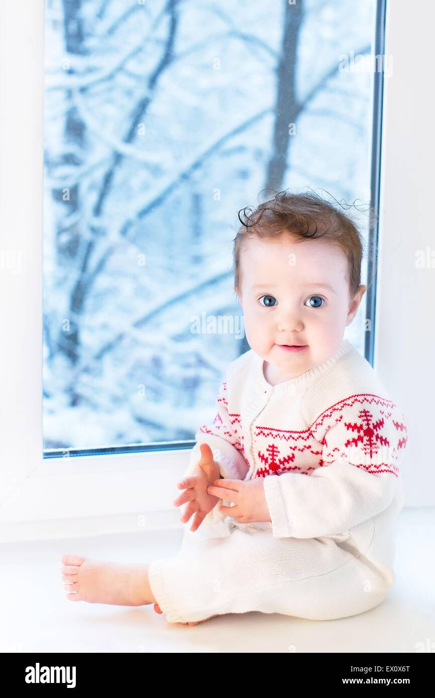 Happy smiling baby sitting at a window watching snow covered trees in ...