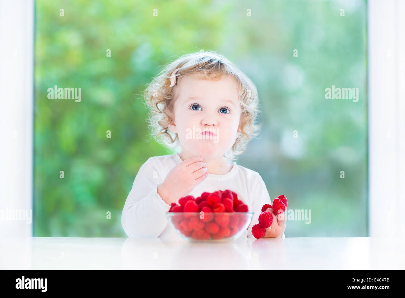 Adorable baby girl eating raspberry at a white table next to a big ...