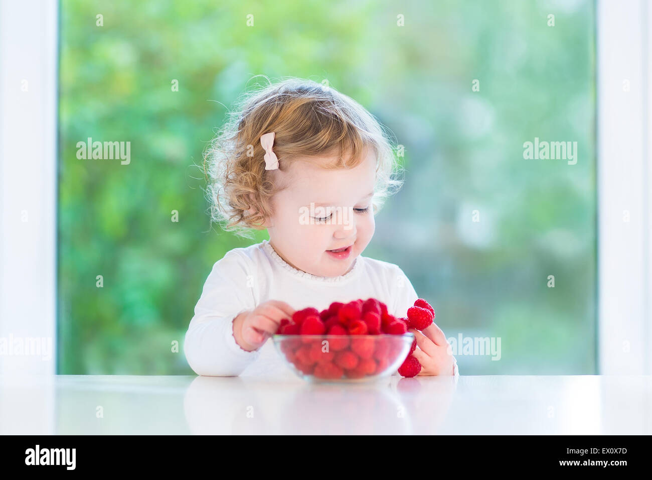Happy smiling baby girl eating raspberry at a white table next to a big ...