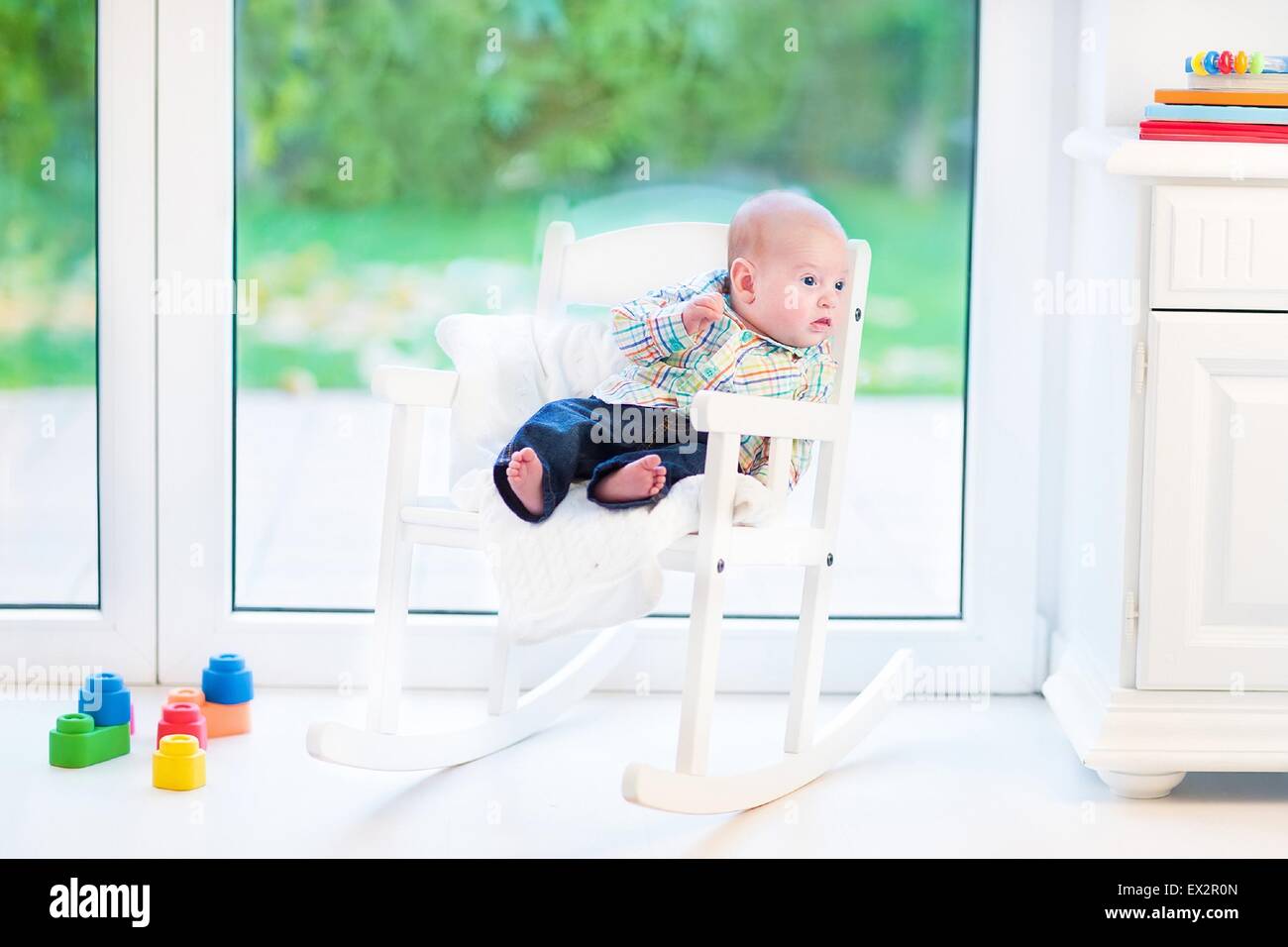 Funny newborn baby boy in a white rocking chair next to a big window ...