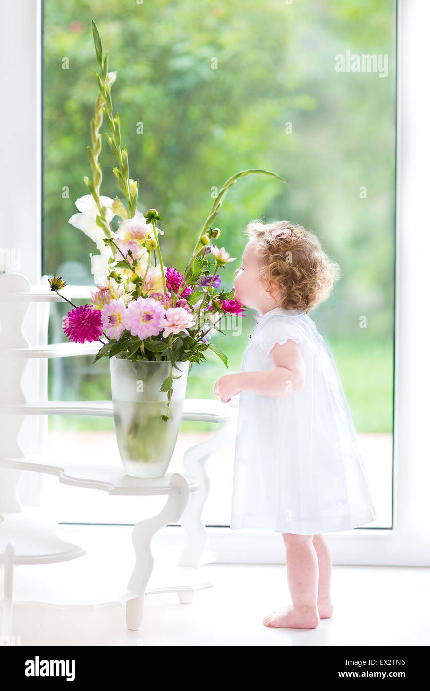 Cute toddler girl smelling fresh flowers at home next to a big window ...