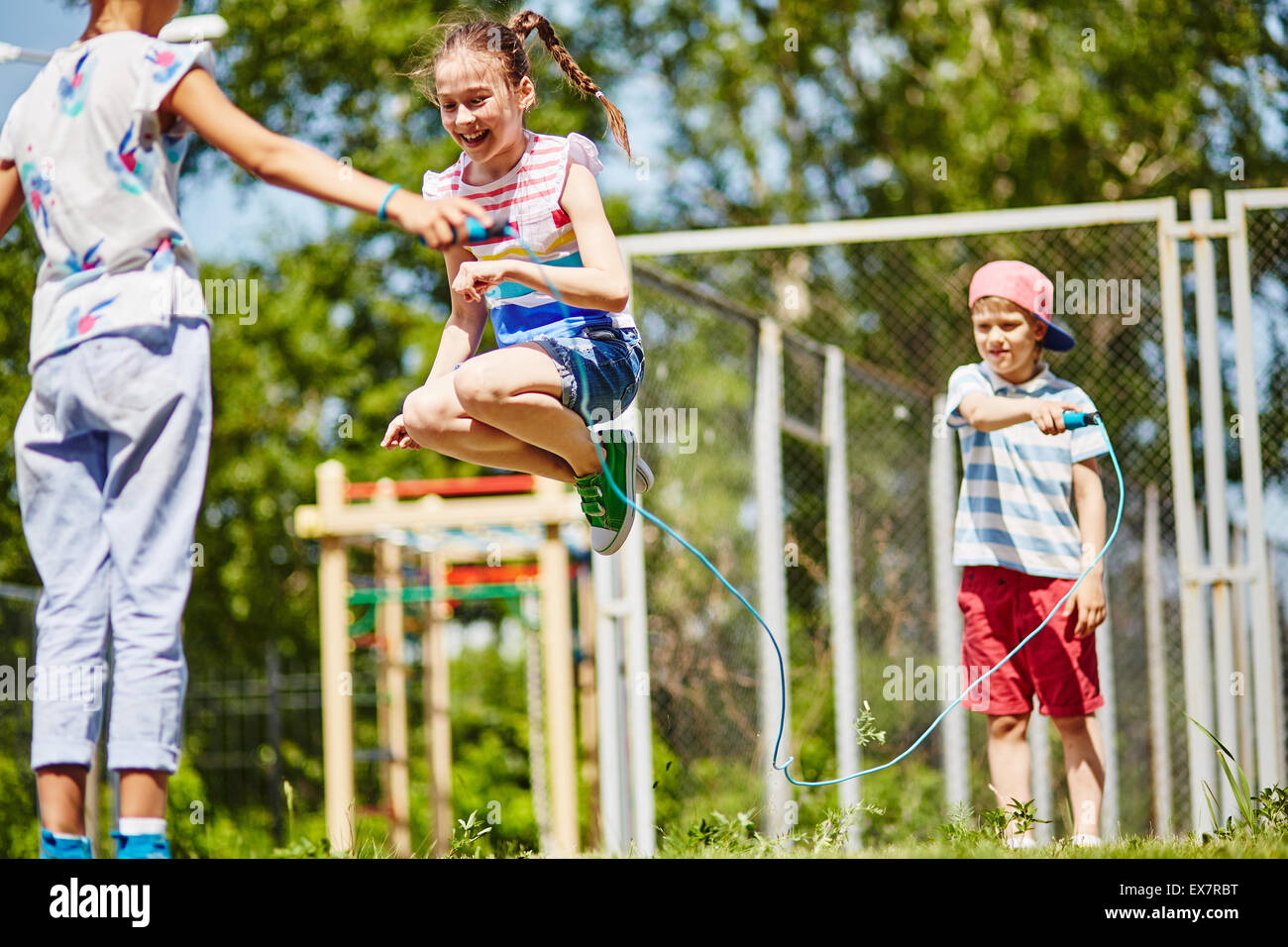 Happy girl jumping over skipping-rope held by her friends outdoors ...