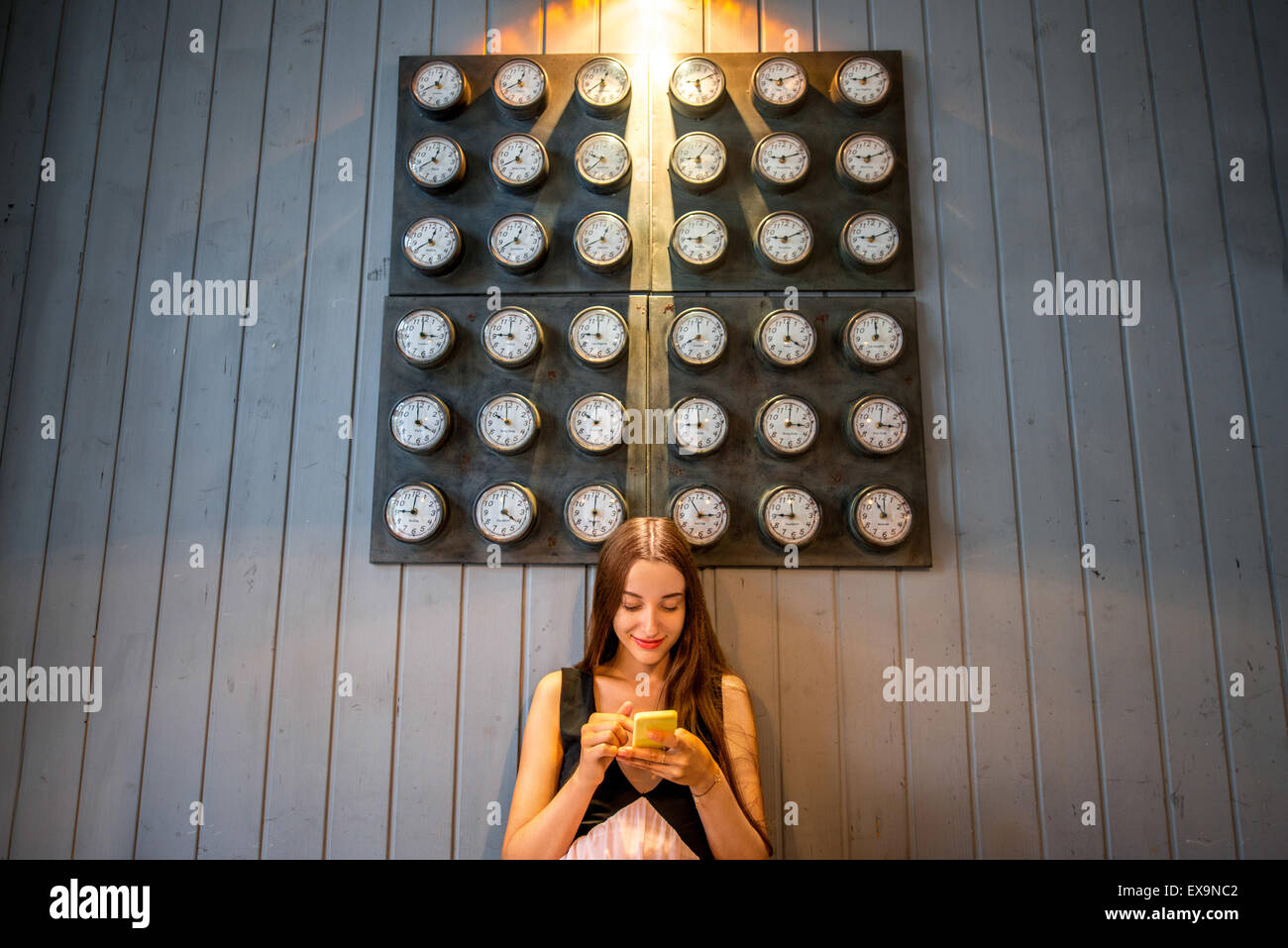 Young woman using mobile phone with many clocks on the wall background ...