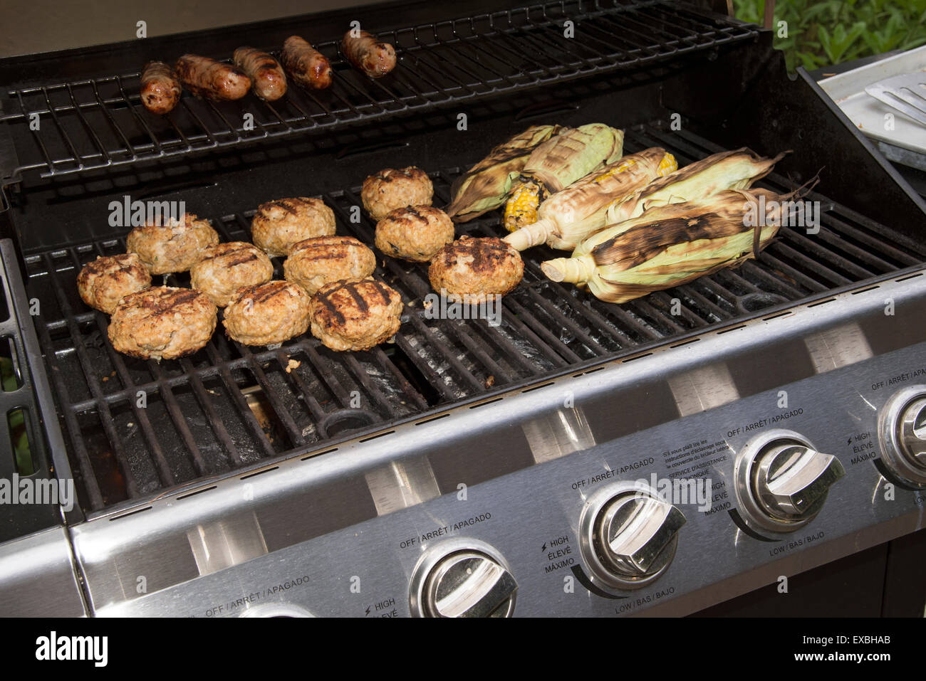 Food cooking on a gas barbecue. Sausages, turkey burgers and corn just ...