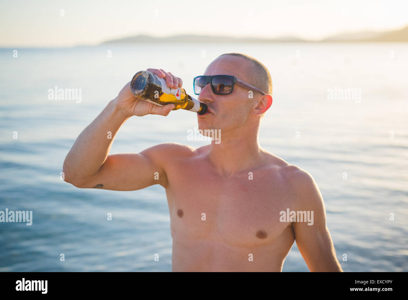 young handsome sporty man drinking beer at the beach sunset in ...