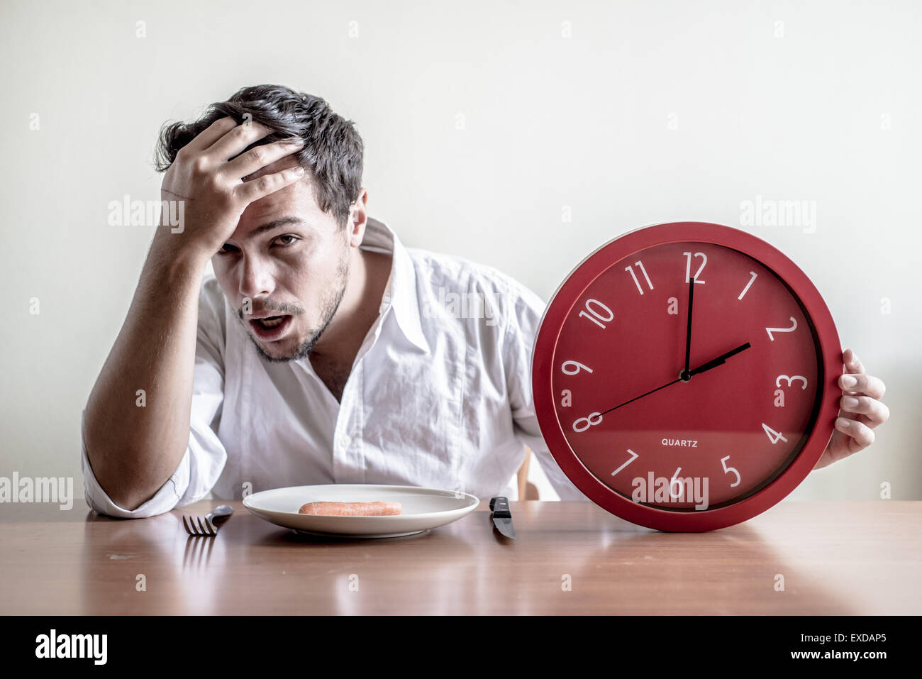 young stylish man with white shirt holding red clock behind a table ...