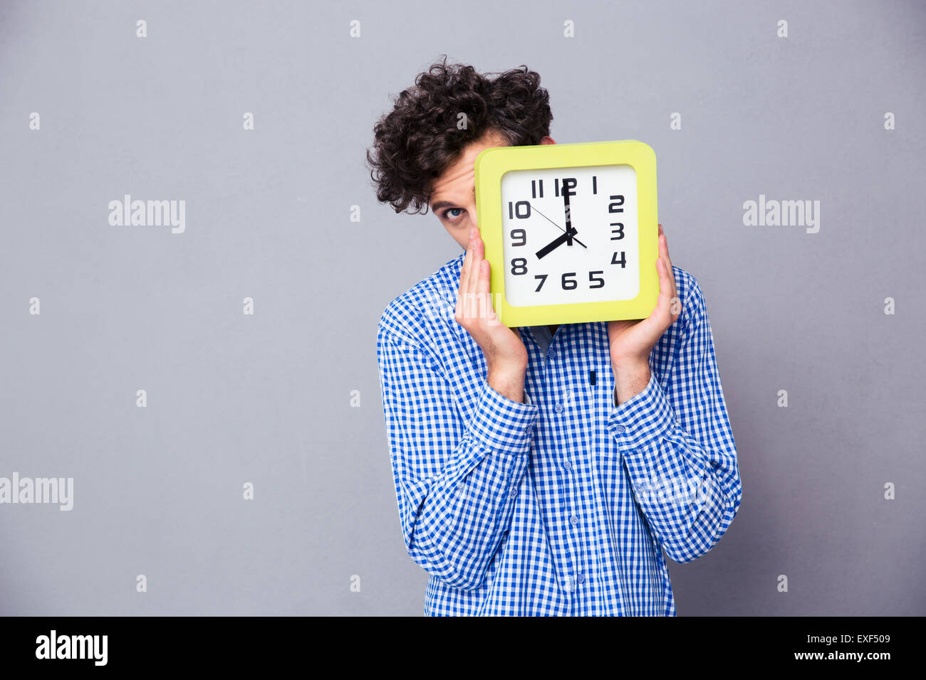 Man covering his face with big clock and looking at camera over gray ...