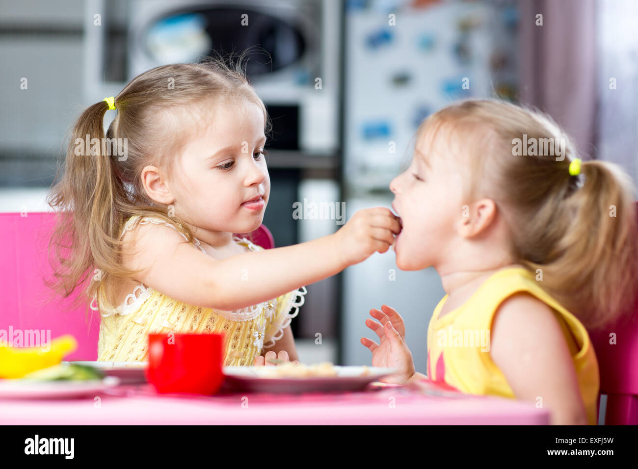 Two little children toddlers eating meal together, one girl feeding ...