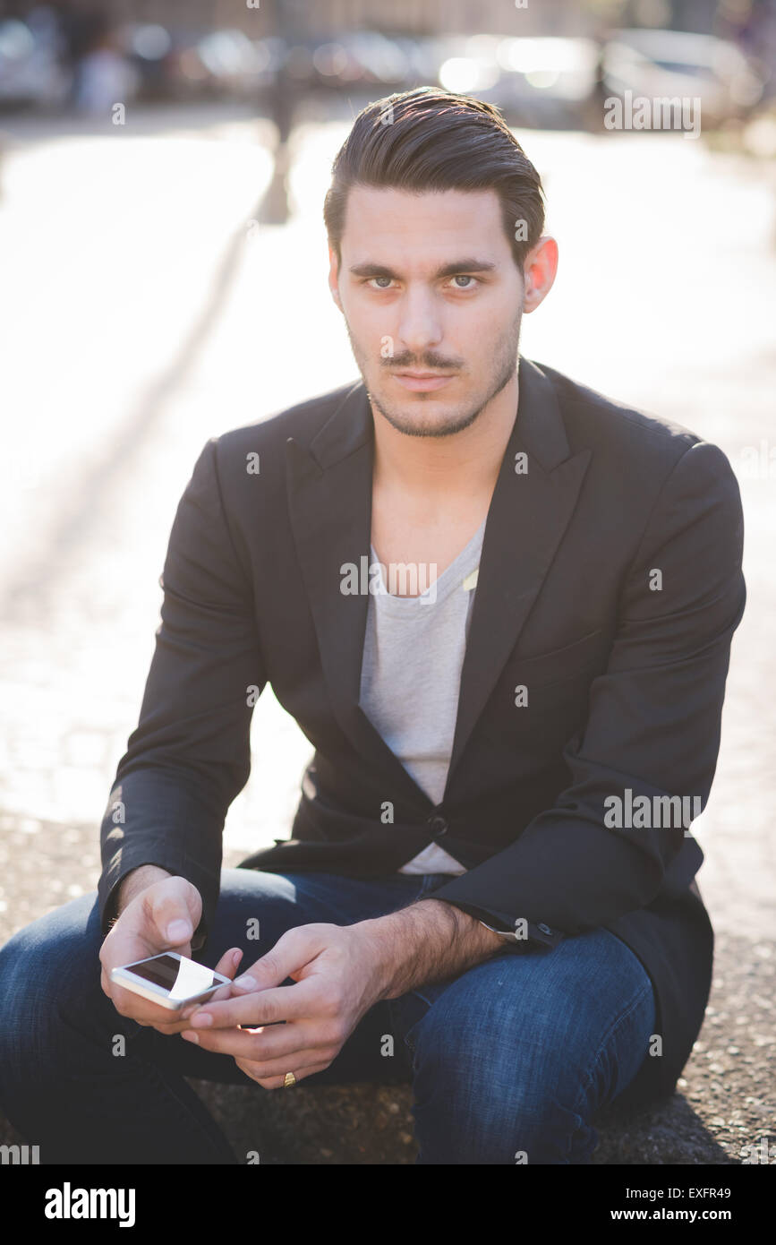 Young handsome italian boy seated on a sidewalk using a smartphone ...