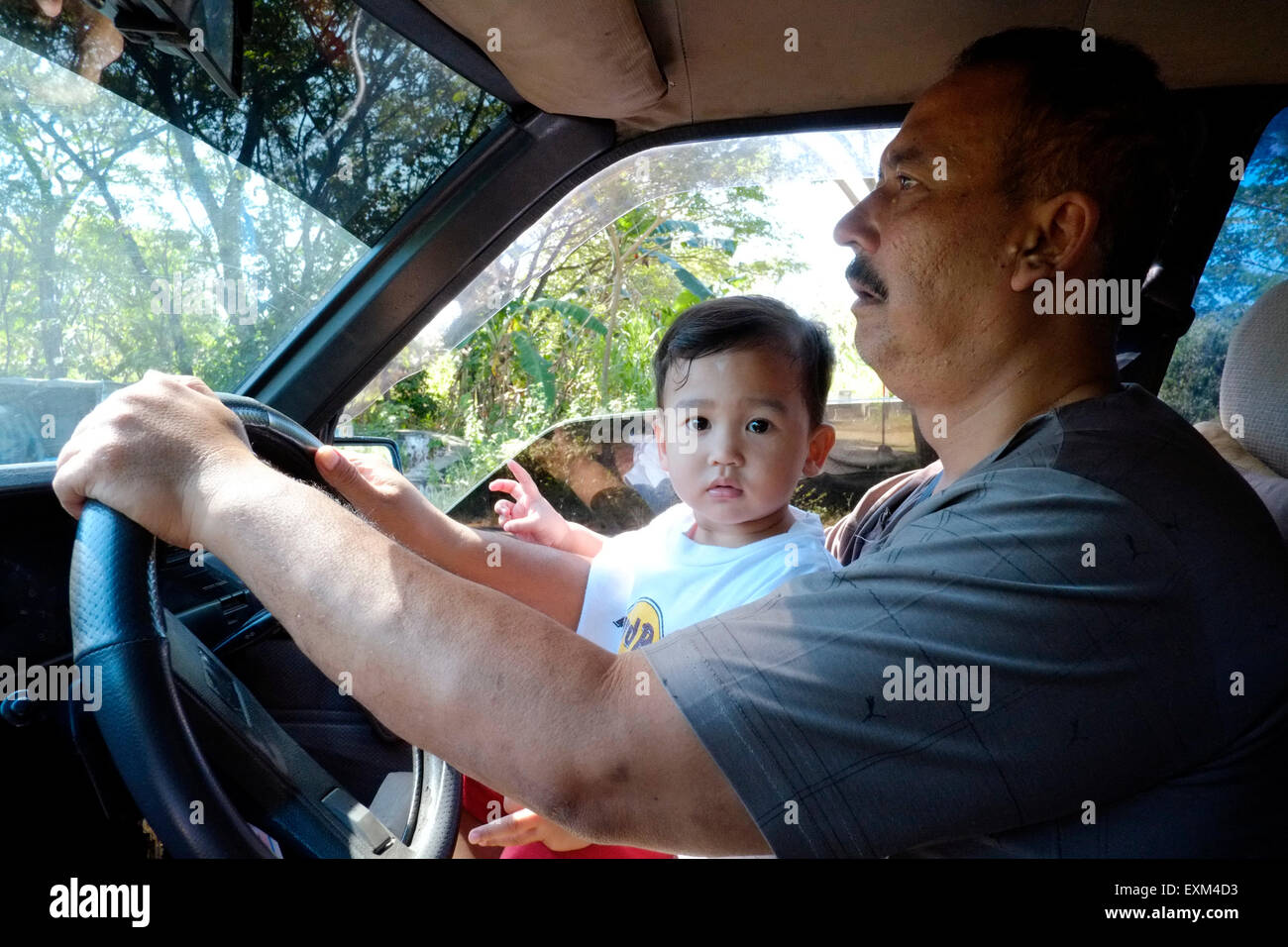 a father driving his car with his young son sitting on his lap in java ...