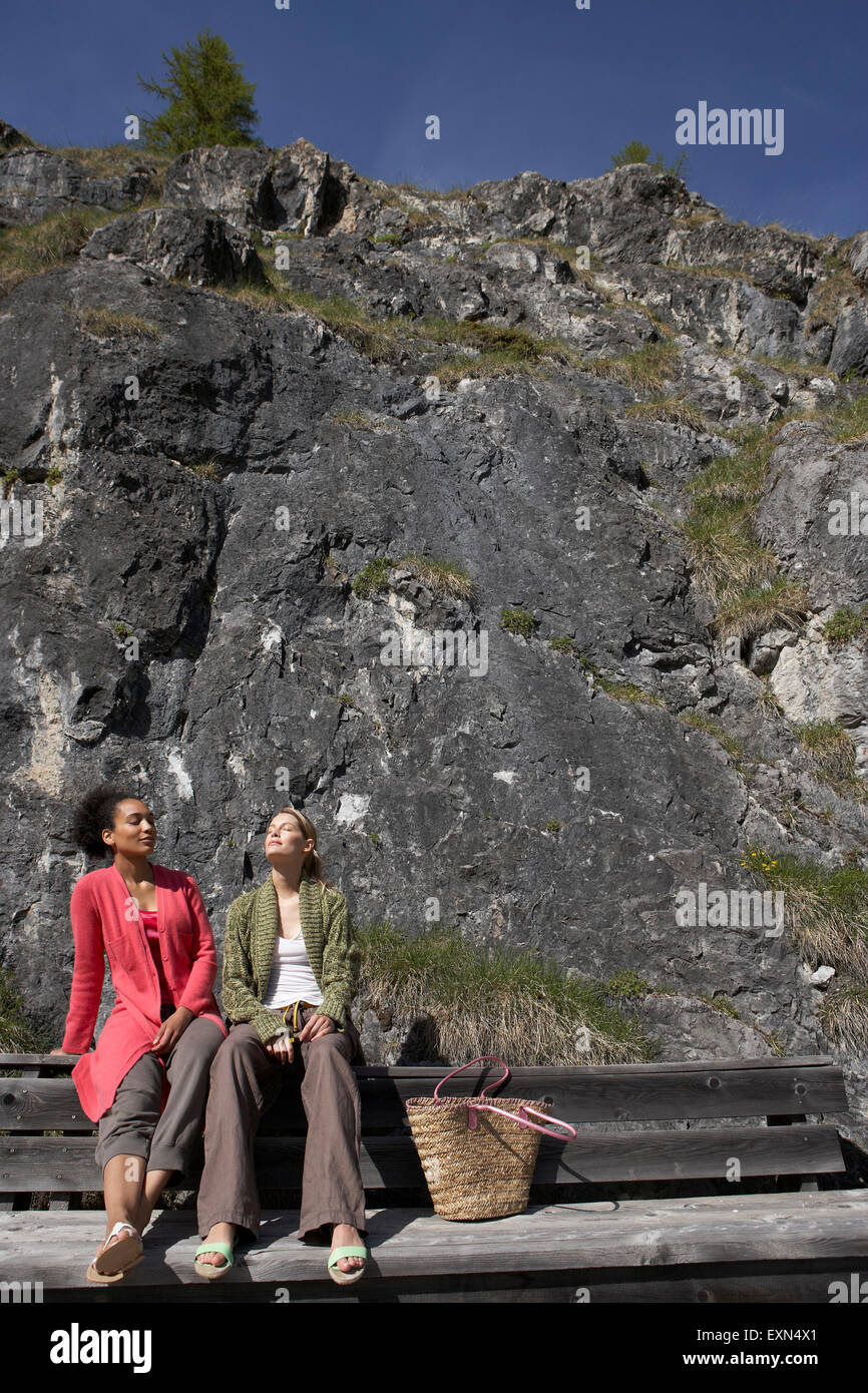 Switzerland, two young women sitting on backrest of a bench sunbathing ...