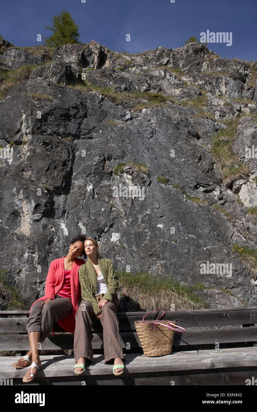 Switzerland, two young women sitting on backrest of a bench sunbathing ...